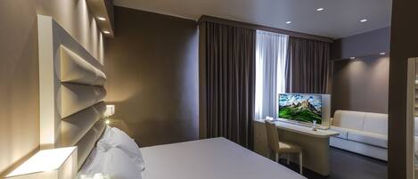 Quarto de hotel moderno com cama, escrivaninha com computador, televisão e sofá.
