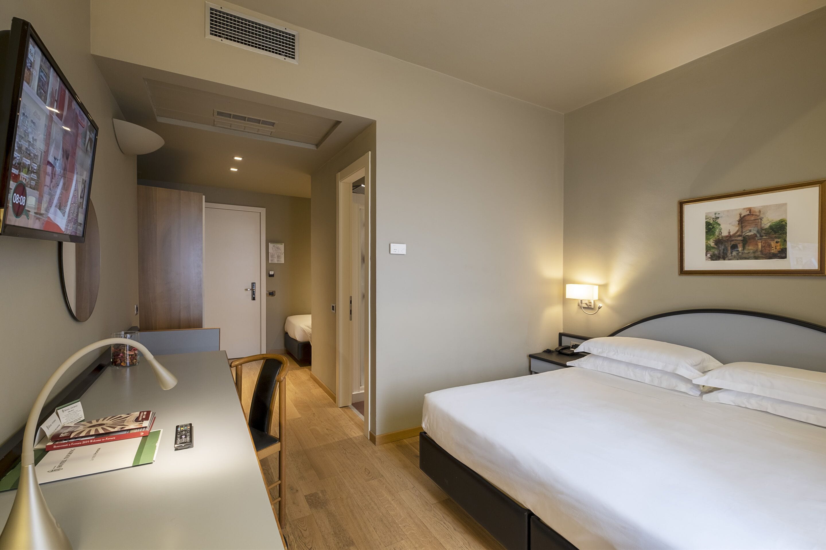 Quarto de hotel com cama, mesa de cabeceira, escrivaninha, cadeira e televisão na parede.