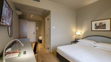 Quarto de hotel com cama, mesa de cabeceira, escrivaninha, cadeira e televisão na parede.