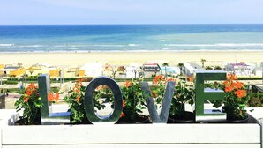 Vista da praia com a palavra 'LOVE' em letras grandes, rodeada por plantas em vasos com flores laranjas.