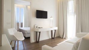 Quarto de hotel moderno com televisão de tela plana, escrivaninha branca, cadeira, sofá e uma grande janela com cortinas.