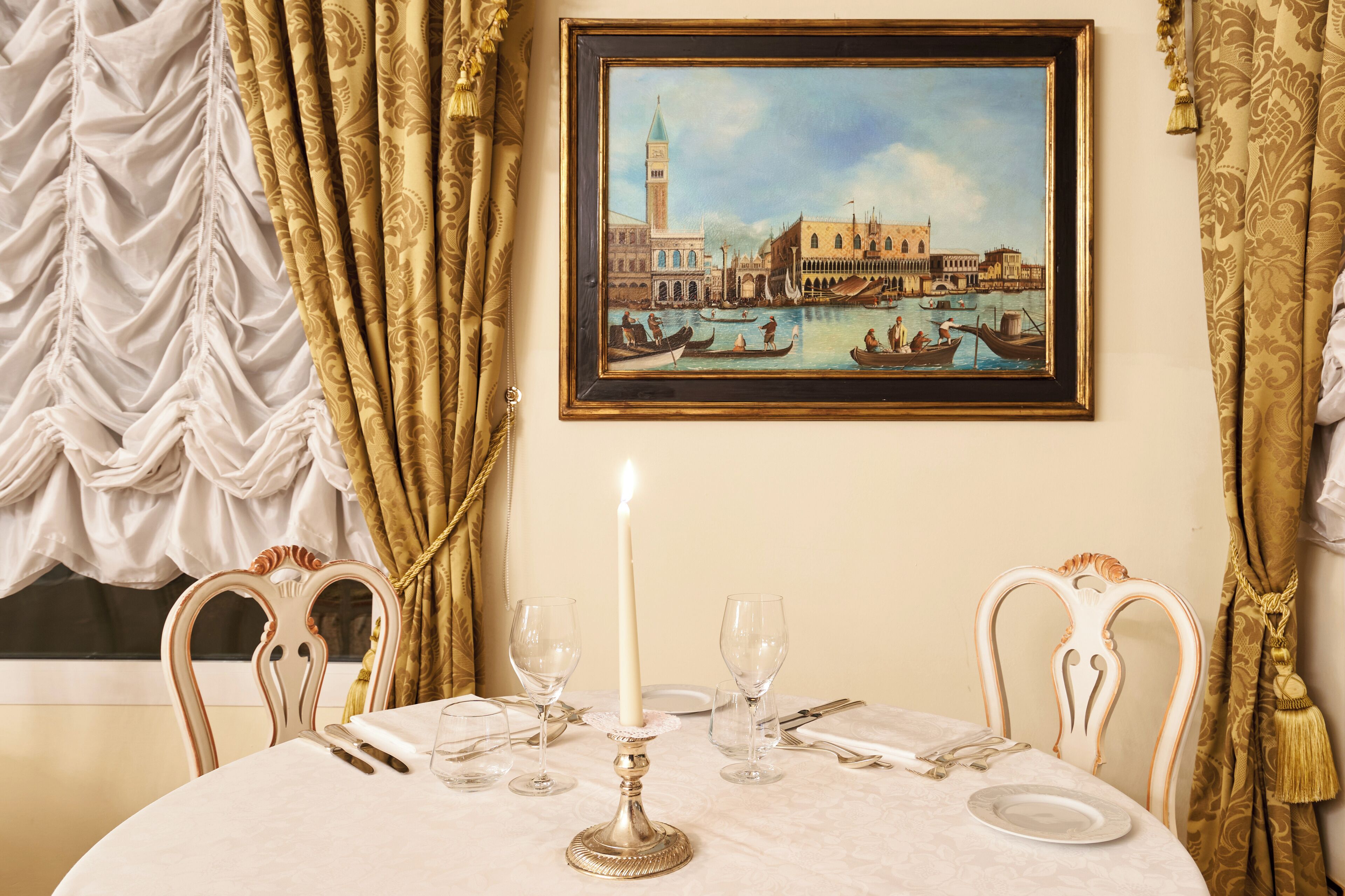Uma mesa de jantar posta com pratos brancos, copos de vinho e talheres, um candelabro aceso e um quadro com um edifício histórico.