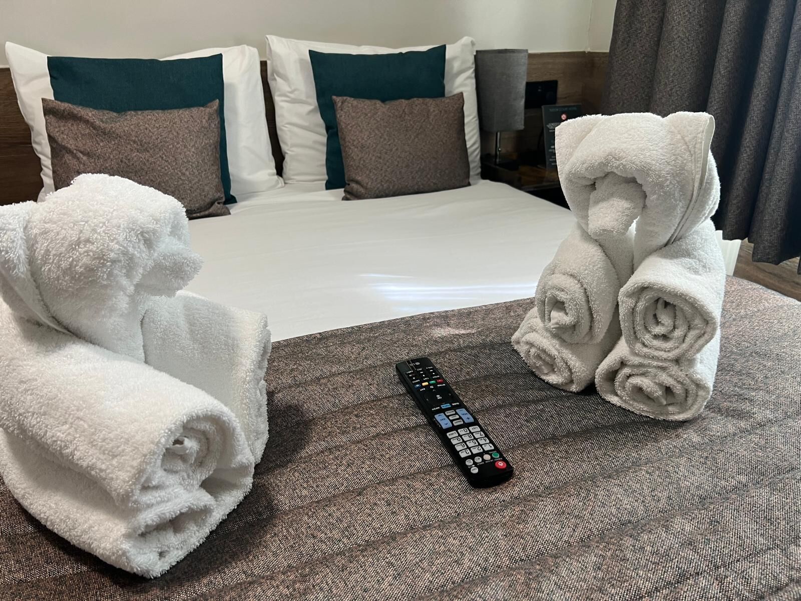 Quarto de hotel com cama bem arrumada, um controle remoto e duas toalhas em formato de ursos sobre a cama.