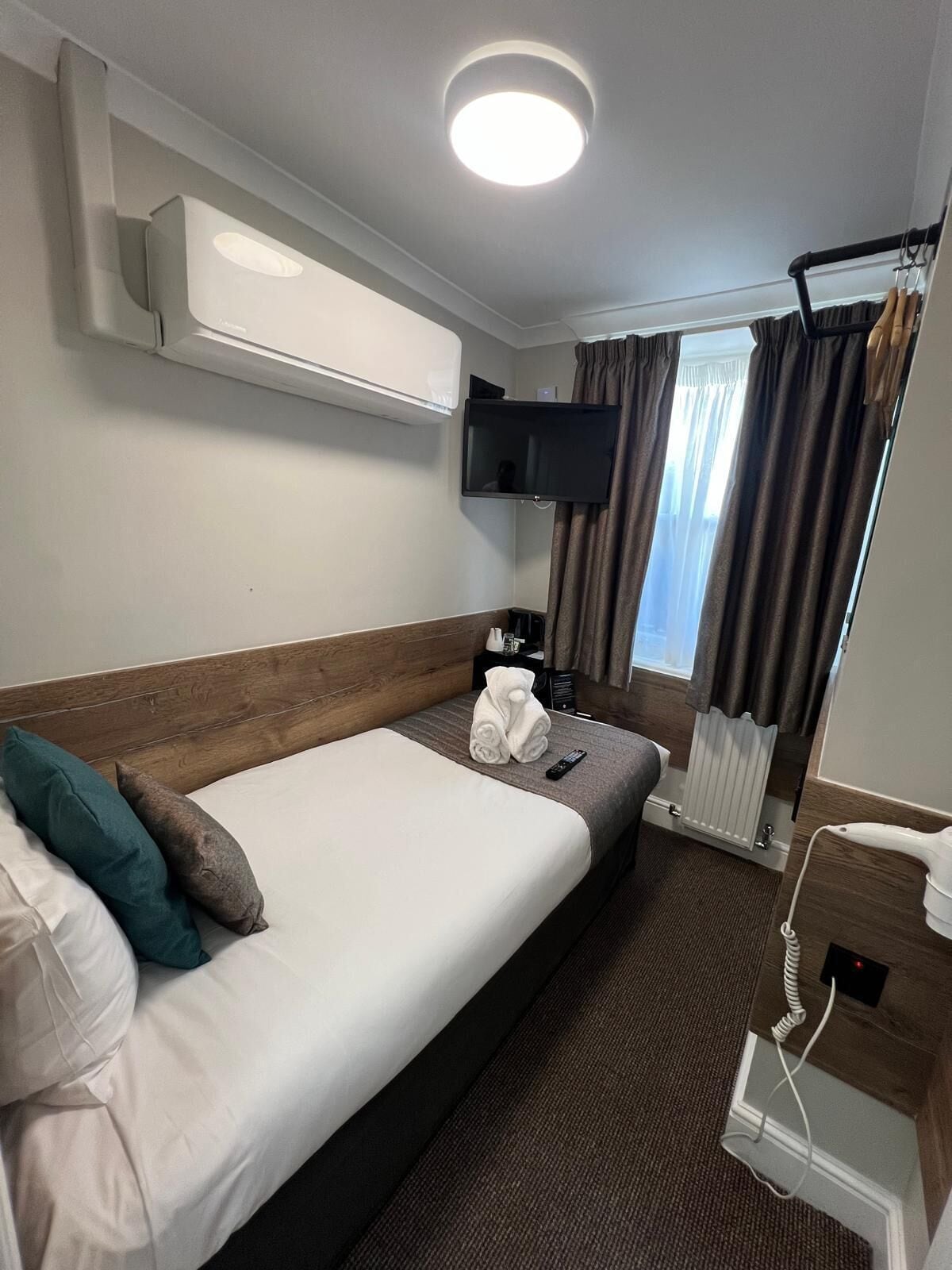 Quarto de hotel com uma cama, televisão fixada na parede, janela com cortinas, radiador e telefone.