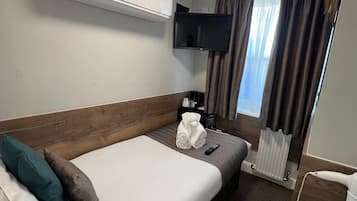 Quarto de hotel com uma cama, televisão fixada na parede, janela com cortinas, radiador e telefone.