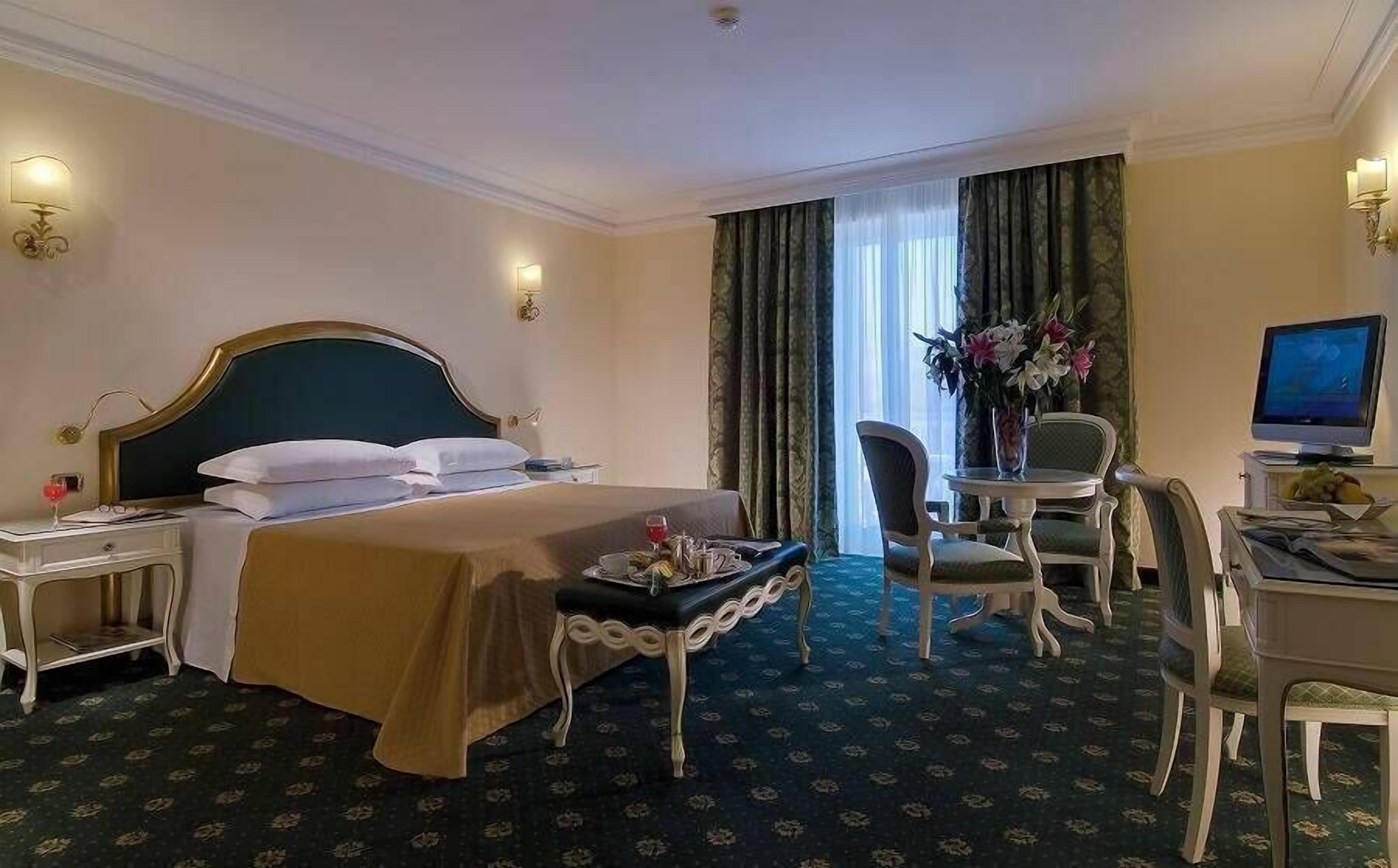 Quarto de hotel com cama, secretária com computador, cadeira, pequena mesa com um vaso de flores e janela com cortinas.