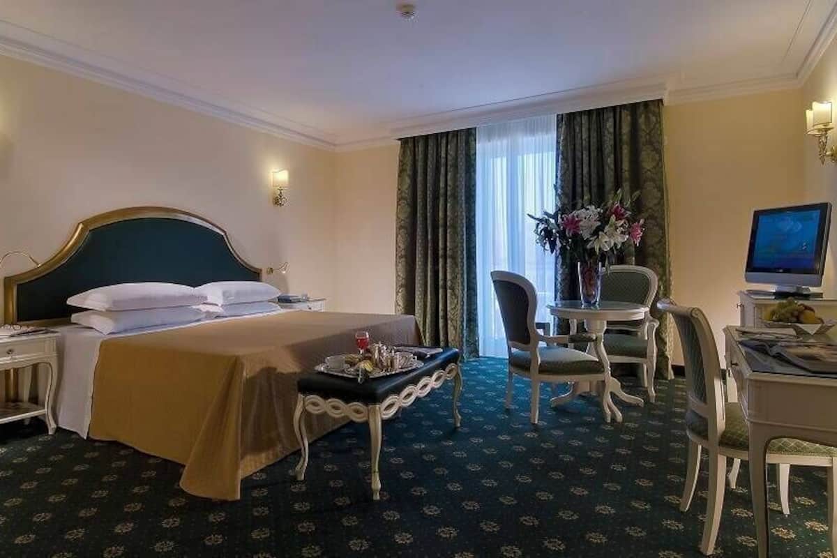 Quarto de hotel com cama, secretária com computador, cadeira, pequena mesa com um vaso de flores e janela com cortinas.