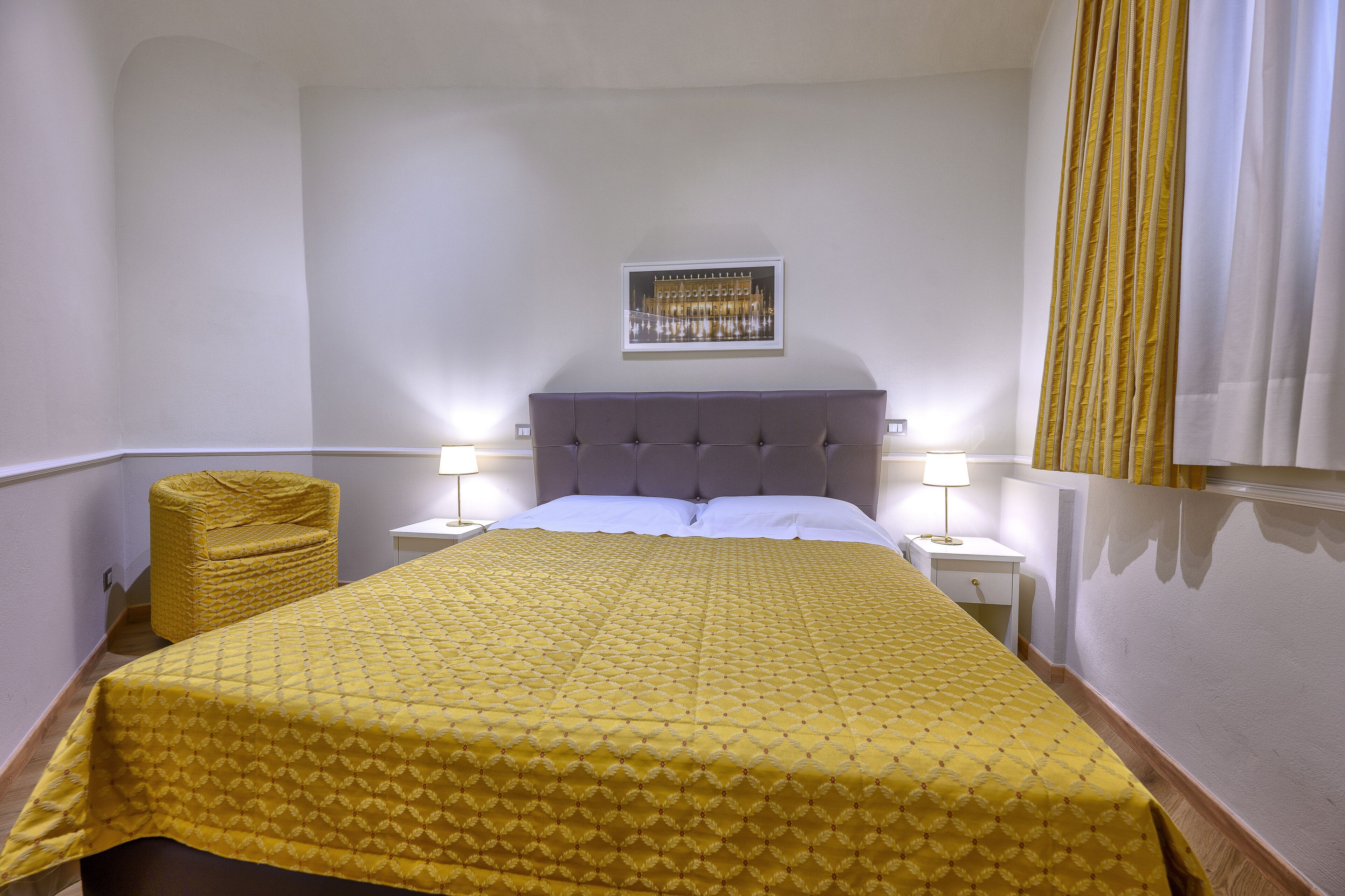 Quarto de hotel com cama grande, cabeceira, mesa de cabeceira, abajur e um quadro pendurado na parede.
