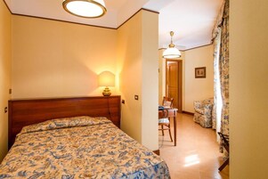 Quarto de hotel com uma cama, uma secretária e uma cadeira. Há uma lâmpada na secretária e uma porta que dá para outro espaço.