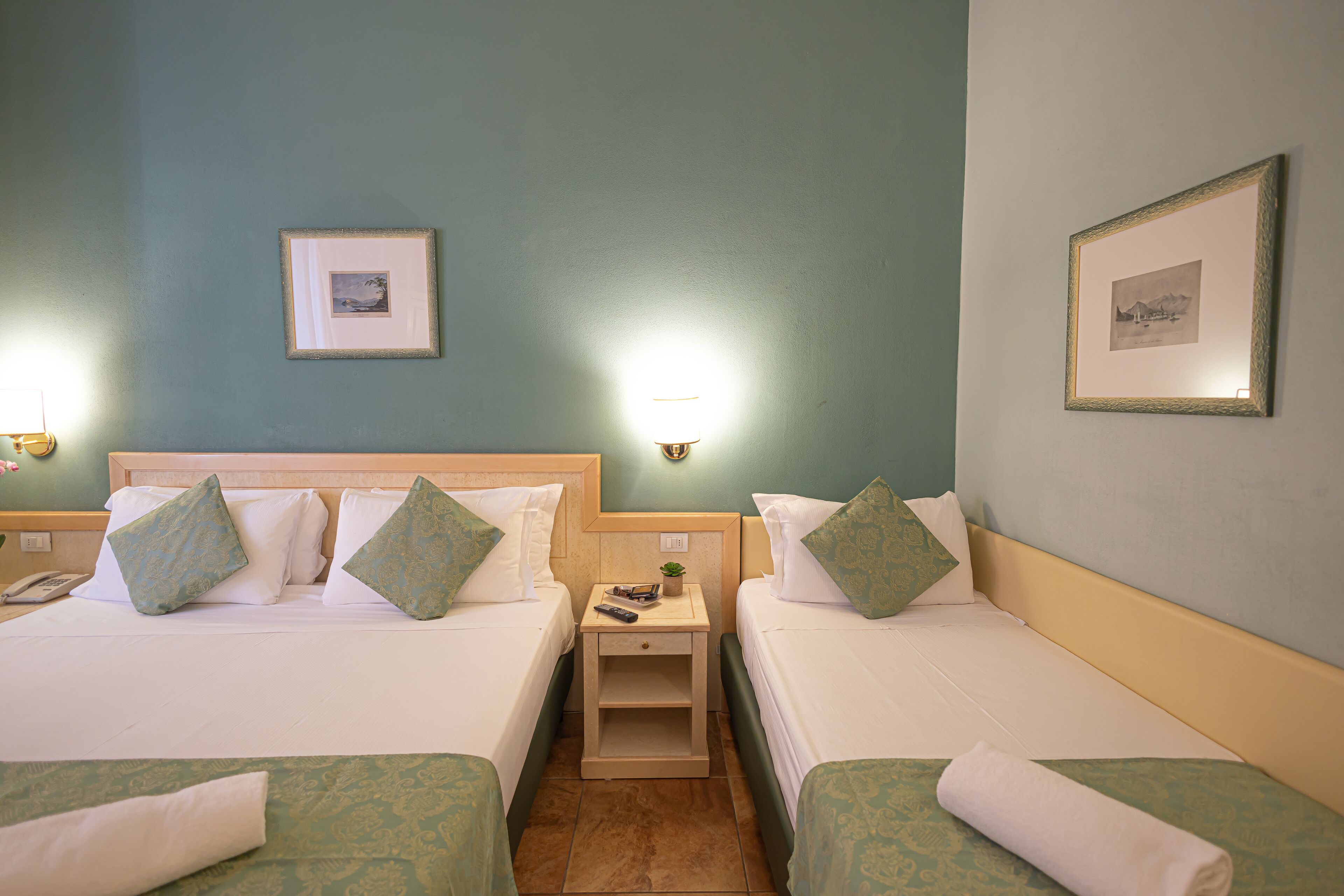Quarto de hotel com duas camas, uma mesa de cabeceira e duas luminárias fixadas na parede.