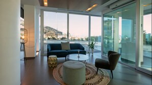 Uma sala de estar moderna com sofá, mesa de centro, banqueta e poltrona, grandes portas de vidro que dão para uma varanda com vista para uma paisagem montanhosa.