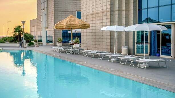 Piscina com espreguiçadeiras e guarda-sóis, cercada por um edifício moderno com janelas amplas e fachada revestida de azulejos.