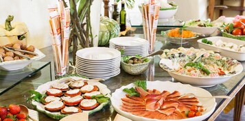 Mesa de buffet com diversos alimentos, incluindo fatias de tomate com queijo, um prato de presunto cru e uma saladeira.