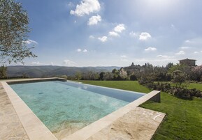 Uma piscina límpida com borda de pedra, rodeada por um relvado bem cuidado e vista para colinas distantes e um edifício histórico.