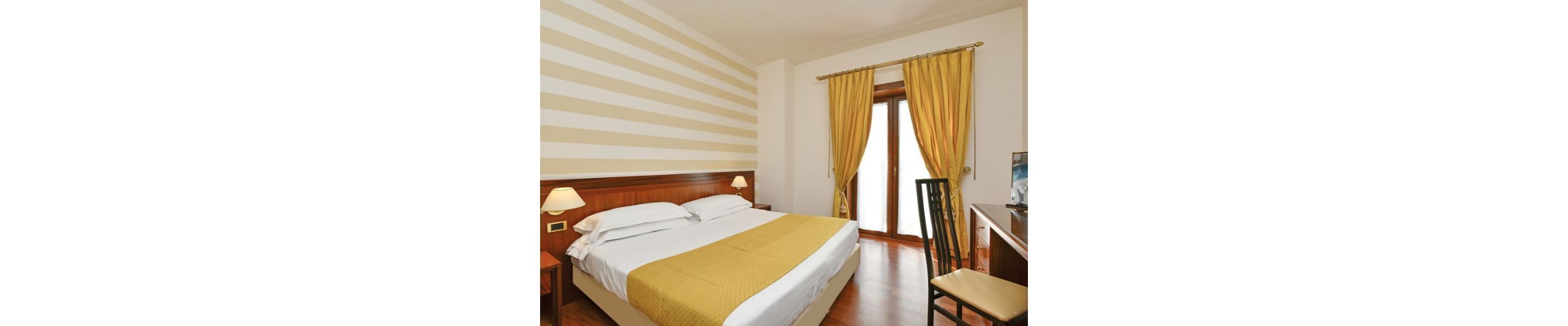 Quarto de hotel com cama, mesa de cabeceira, cadeira e janela com cortinas.