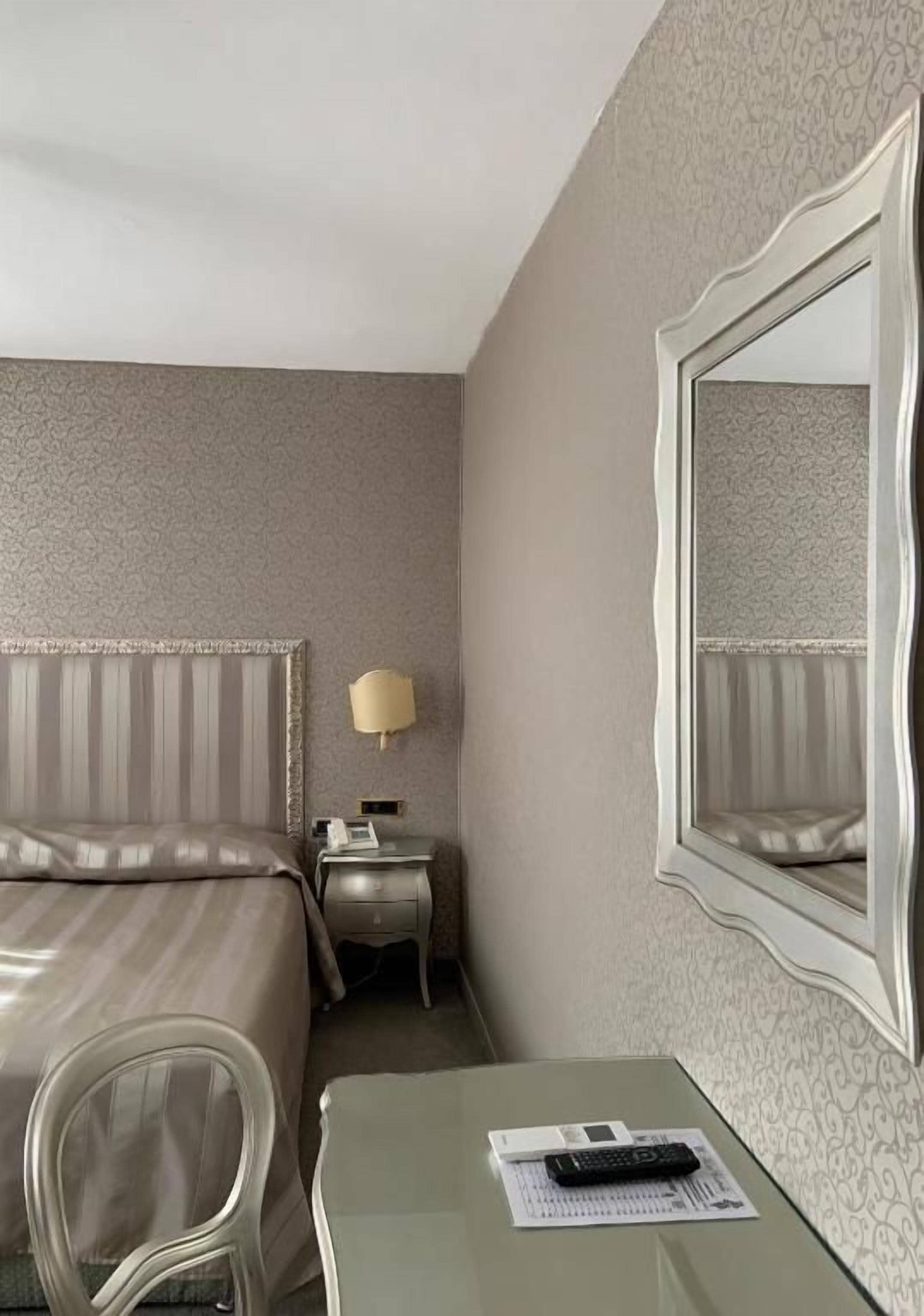 Quarto de hotel com cama, escrivaninha, cadeira, mesa de cabeceira, luminária e espelho.
