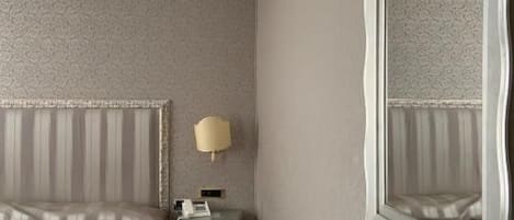 Quarto de hotel com cama, escrivaninha, cadeira, mesa de cabeceira, luminária e espelho.