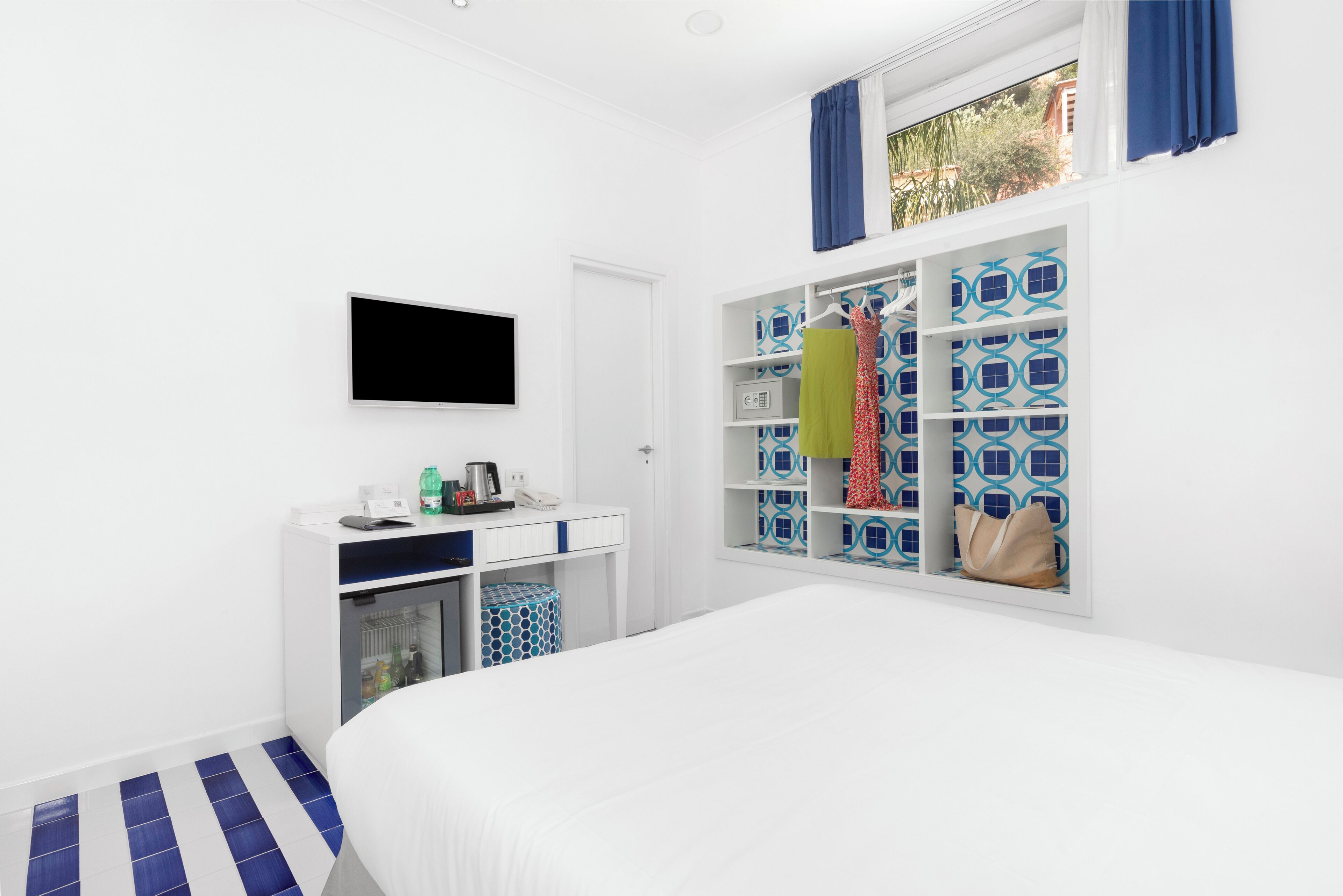 Quarto de hotel com uma cama, uma escrivaninha com televisão de tela plana, um guarda-roupa com prateleiras estampadas e uma janela com cortinas.