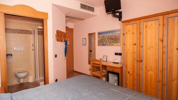 Quarto de hotel com cama, guarda-roupa de madeira, escrivaninha com cadeira, chuveiro e sanita.