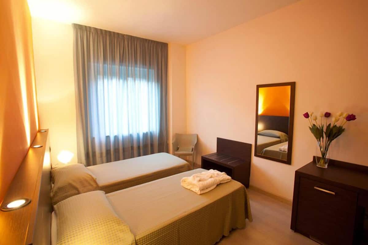 Quarto de hotel com duas camas, uma escrivaninha, uma cadeira, um aparador, um espelho e uma janela com cortinas.