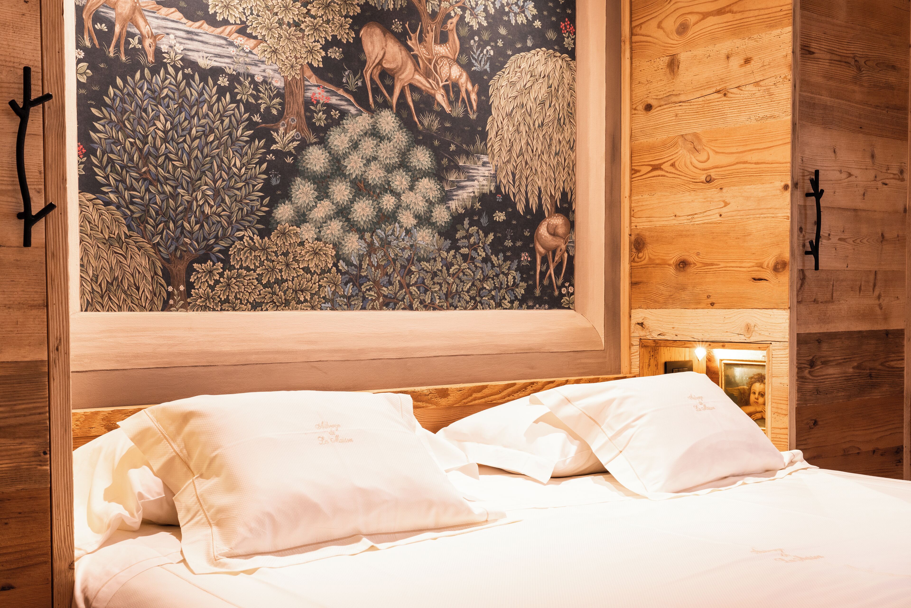 Quarto de cabana de madeira com cama, roupa de cama branca e uma grande pintura de veados e árvores na parede.