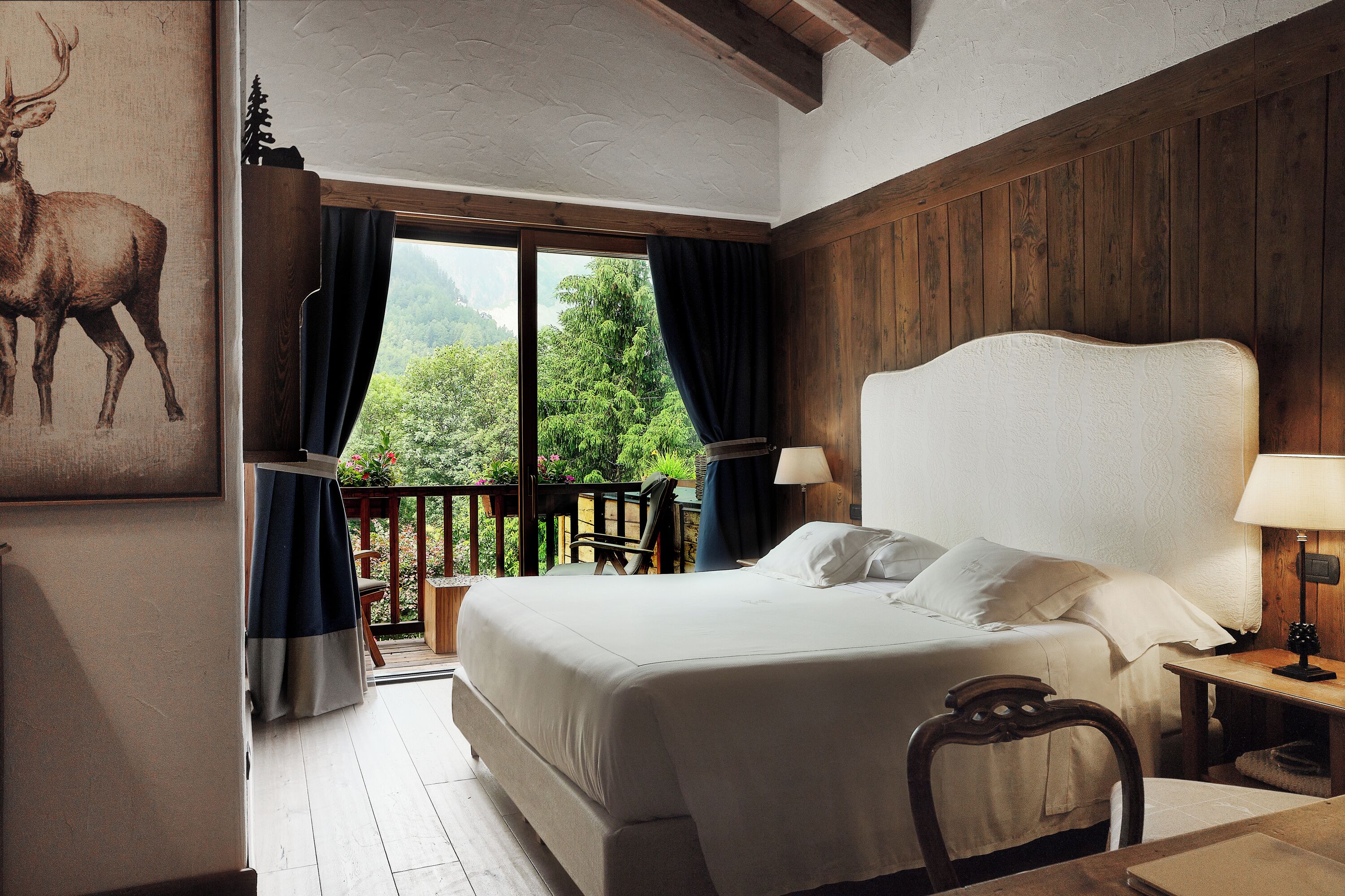 Quarto com uma cama grande, cabeceira de madeira, mesa de cabeceira com abajur e vista para o verde pela janela.