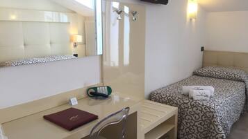 Quarto de hotel com uma cama, uma escrivaninha, uma cadeira, um espelho e uma TV na parede.
