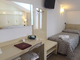 Quarto de hotel com uma cama, uma escrivaninha, uma cadeira, um espelho e uma TV na parede.