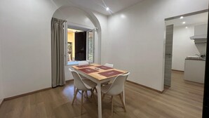 Uma área de refeições com mesa e cadeiras, uma janela com cortinas, e uma cozinha ao fundo.