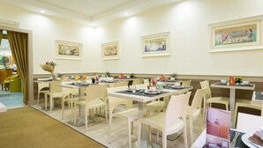 Uma sala de jantar de hotel com várias mesas e cadeiras, obras de arte emolduradas nas paredes e chão coberto de carpete.
