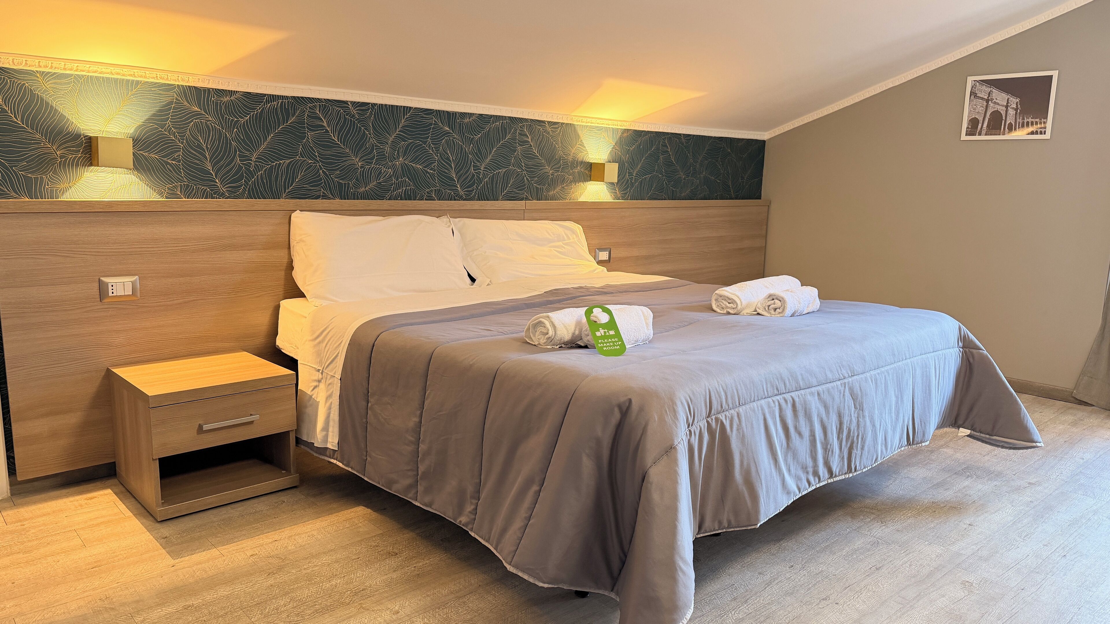 Cama arrumada com um edredom cinzento e duas toalhas enroladas, mesa de cabeceira de madeira com gaveta e luminária fixa na parede.