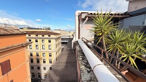 Uma rua estreita com edifícios de ambos os lados, uma varanda visível com uma planta e céu claro.