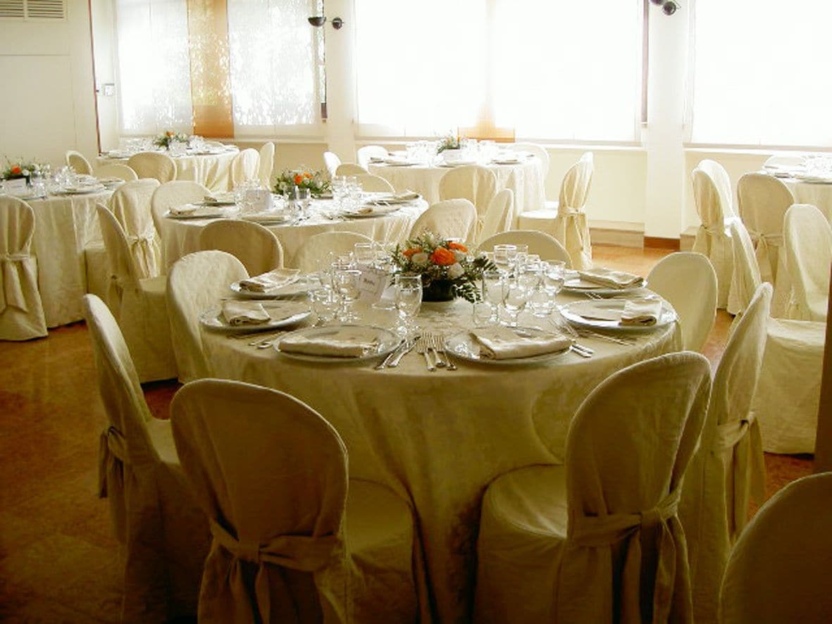 Um salão de festas com mesas redondas preparadas para um evento formal, cada uma com toalhas brancas, cadeiras e centros de mesa.