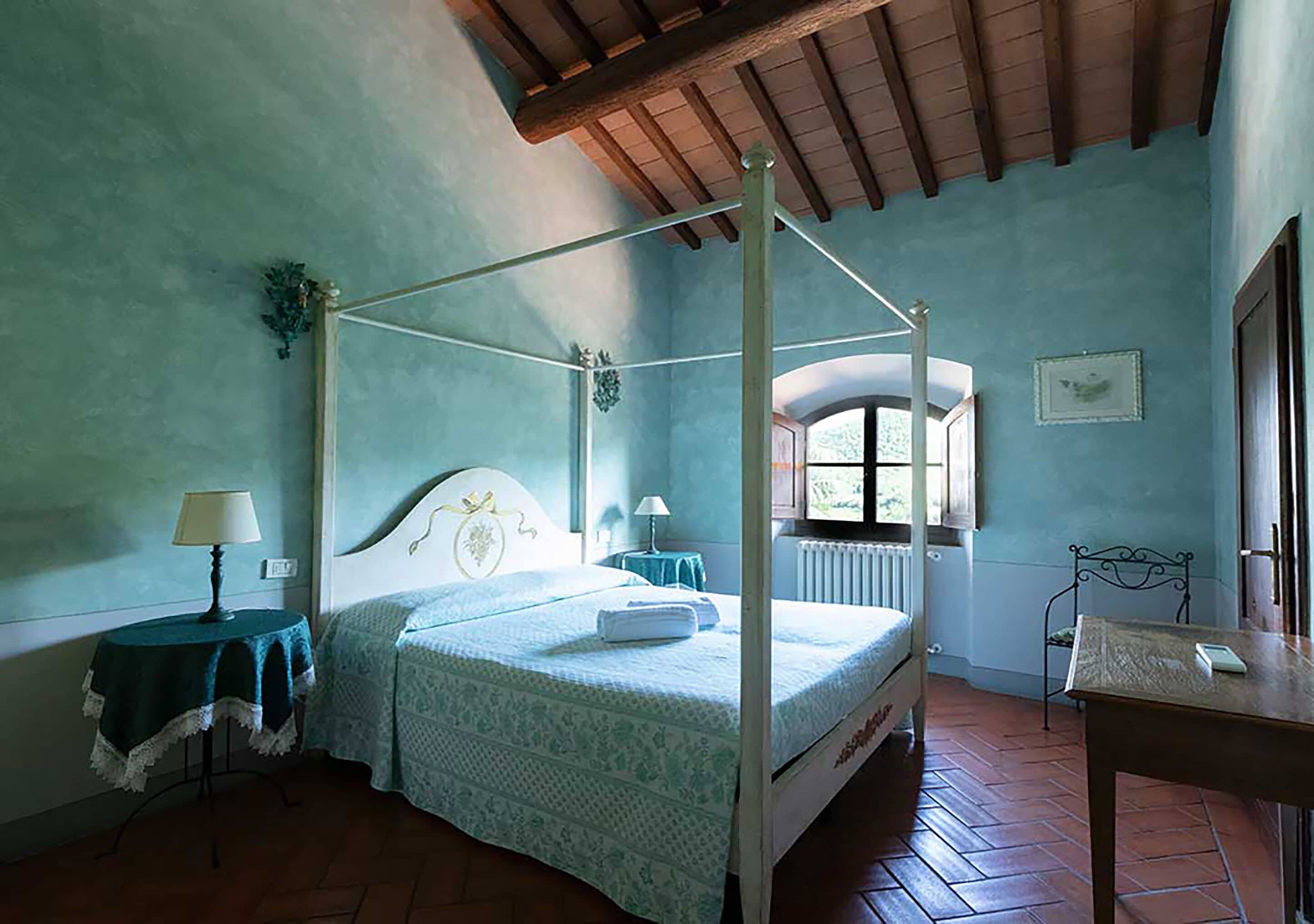 Quarto com uma cama de dossel, uma mesinha de cabeceira com abajur, uma janela com vista e um radiador.