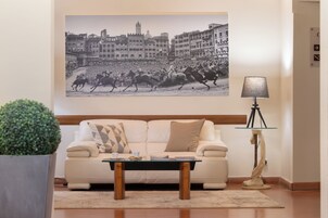 Uma sala de estar com um sofá bege, uma mesa de centro de vidro, um abajur e uma fotografia grande em preto e branco na parede.