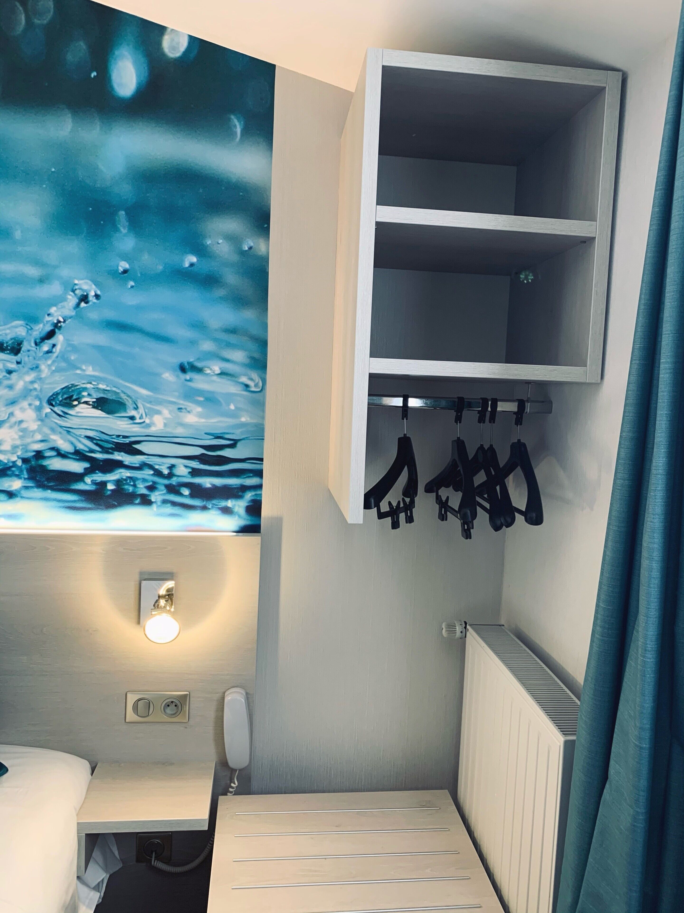 Quarto pequeno com uma cama, prateleira na parede, radiador e uma obra de arte com tema aquático.