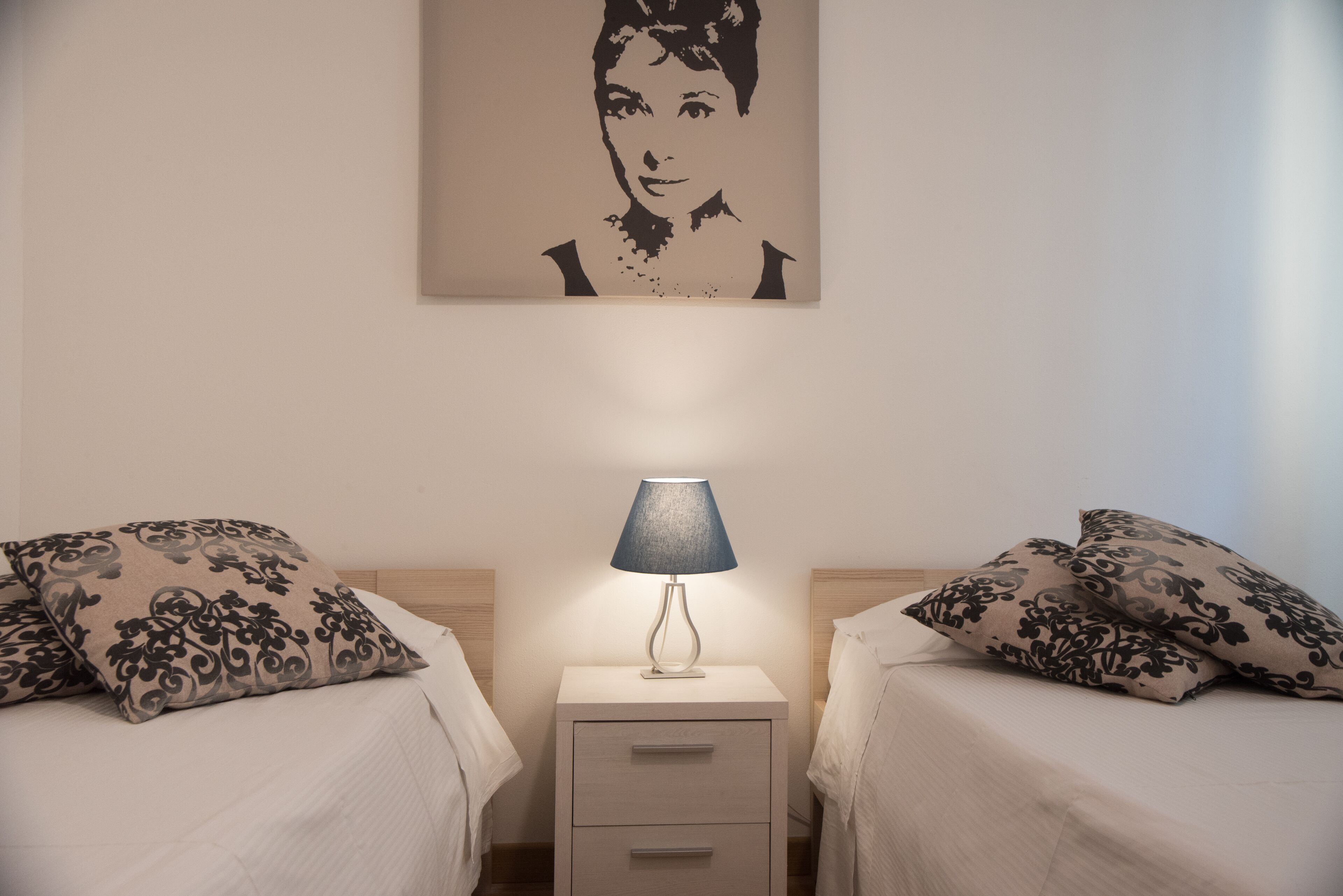 Quarto com duas camas, cada uma com almofadas estampadas, mesa de cabeceira com abajur e obra de arte na parede.