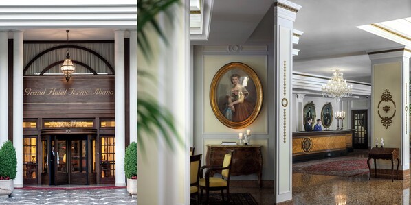 Entrada imponente de um hotel, com uma porta giratória, balcão de recepção e um candelabro.