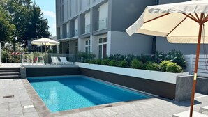 Uma piscina exterior moderna, com água azul translúcida, rodeada por espreguiçadeiras brancas e guarda-sóis bege.