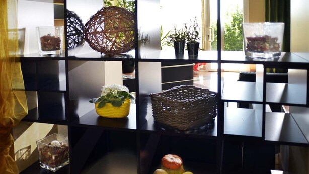 Uma estante preta com diversos objetos decorativos, incluindo uma cesta trançada, uma tigela de vidro com flores secas e uma abóbora pequena.
