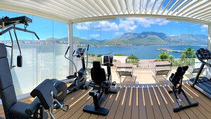 Academia com equipamentos de exercício e vista para uma cidade costeira e montanhas.