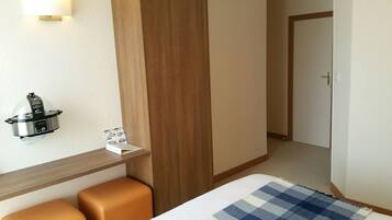 Quarto de hotel com escrivaninha de madeira, um pequeno espaço de estar, cama com colcha xadrez e chaleira fixada na parede.