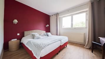Cama de casal com cabeceira vermelha e roupa de cama branca, luminária de cabeceira, cesto de vime e janela com cortinas.