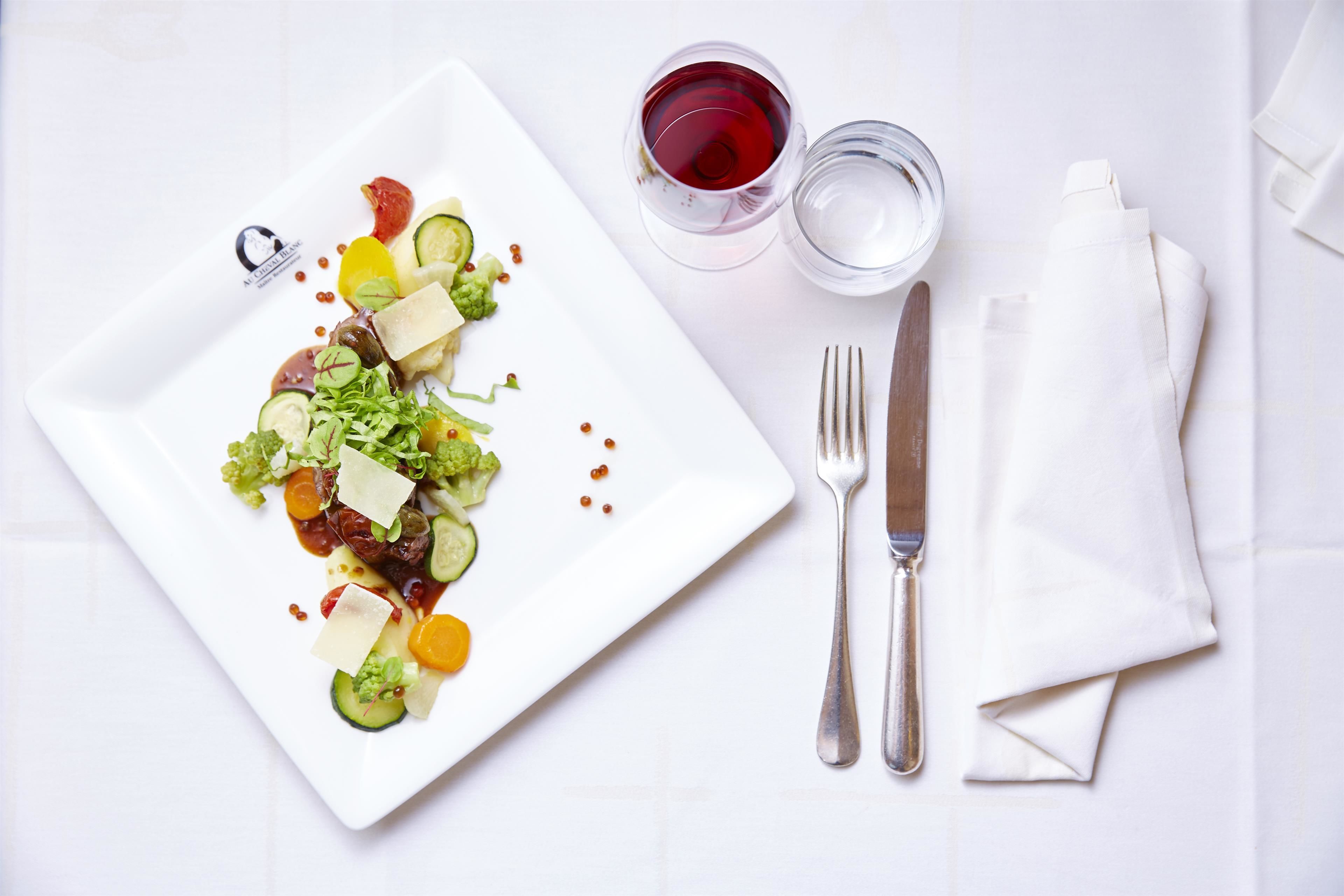 Um prato branco com salada, um copo de vinho tinto e uma toalha dobrada com talheres sobre uma mesa branca.