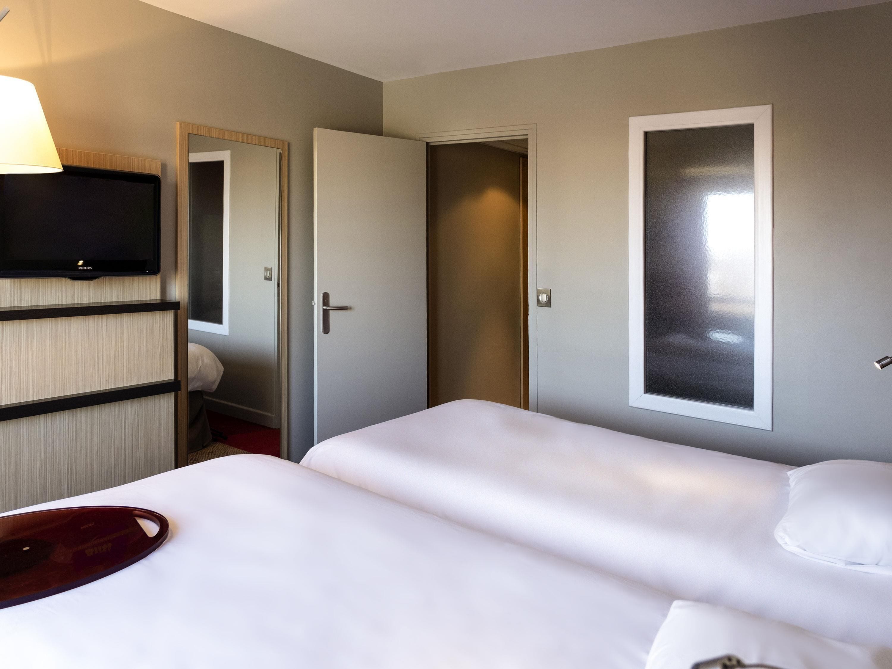Quarto de hotel com duas camas, televisão e porta de acesso a outro espaço.