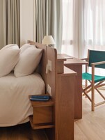 Quarto com cama, mesa de cabeceira, luminária, cadeira e pequena estante de madeira com livros.
