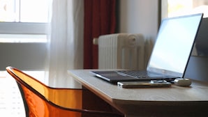 Um laptop sobre uma mesa de madeira, com uma cadeira, um radiador e uma janela com cortinas.