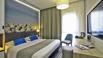 Quarto de hotel com cama, escrivaninha, cadeira, luminária e uma janela grande.