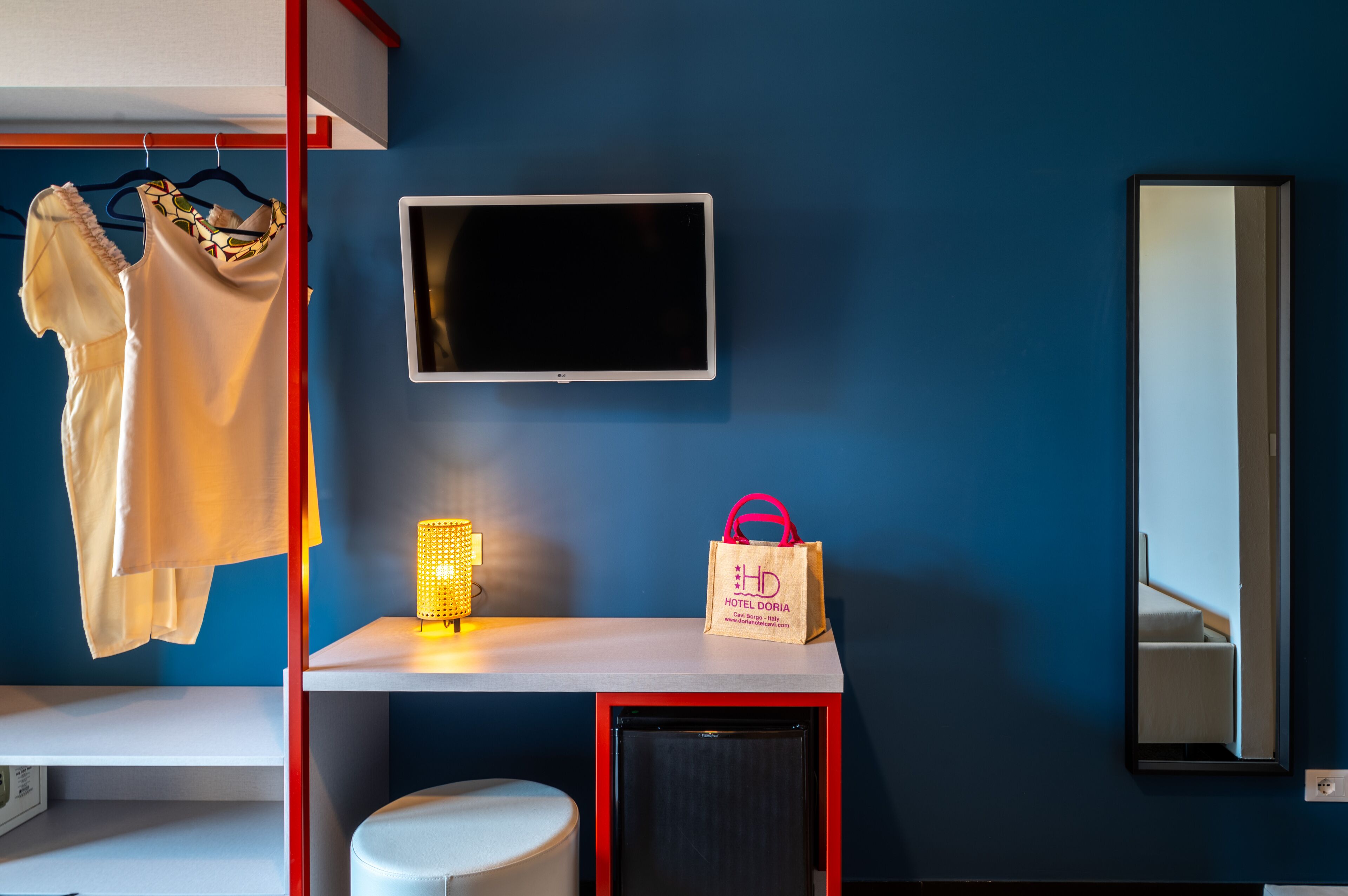 Quarto de hotel com televisão, uma pequena mesa com abajur, espelho e cabides.
