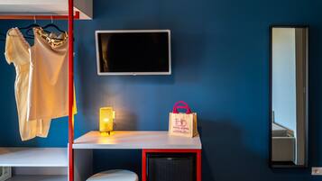 Quarto de hotel com televisão, uma pequena mesa com abajur, espelho e cabides.